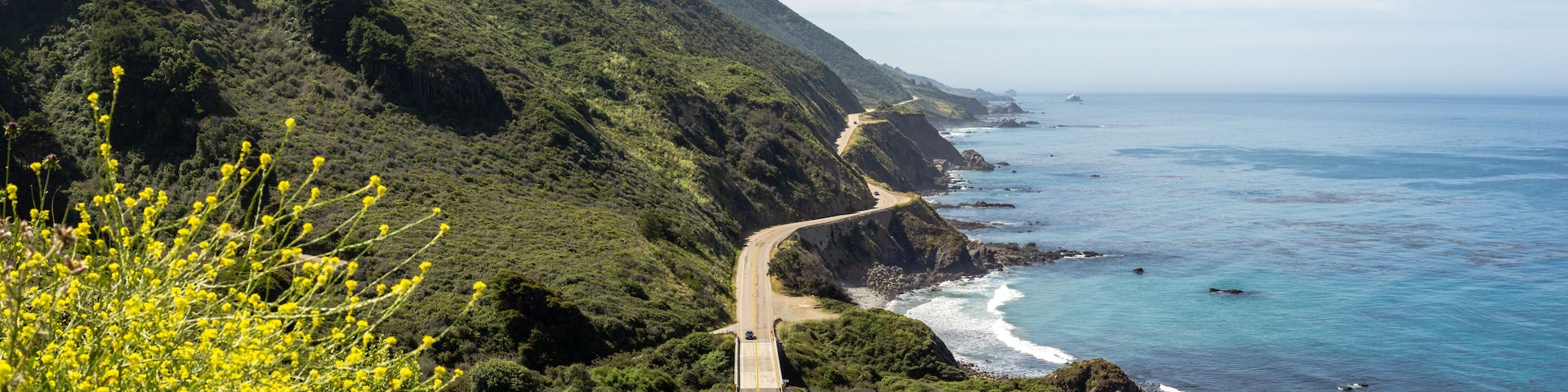 Uma estrada costeira serpenteia por uma paisagem montanhosa, com vista para o oceano.