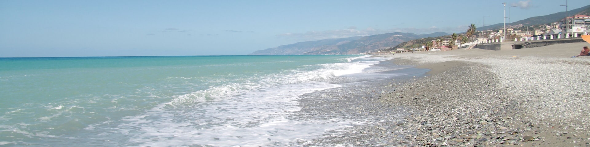 Uma praia com águas turquesa cristalinas, areal de pedras e edifícios ao fundo.