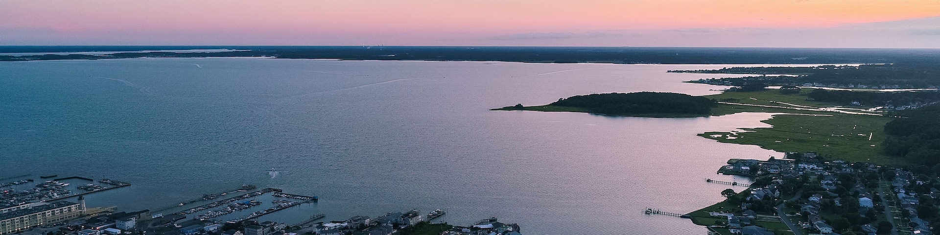Vista aérea de uma cidade costeira ao pôr do sol, com uma grande massa de água, um cais e áreas residenciais.