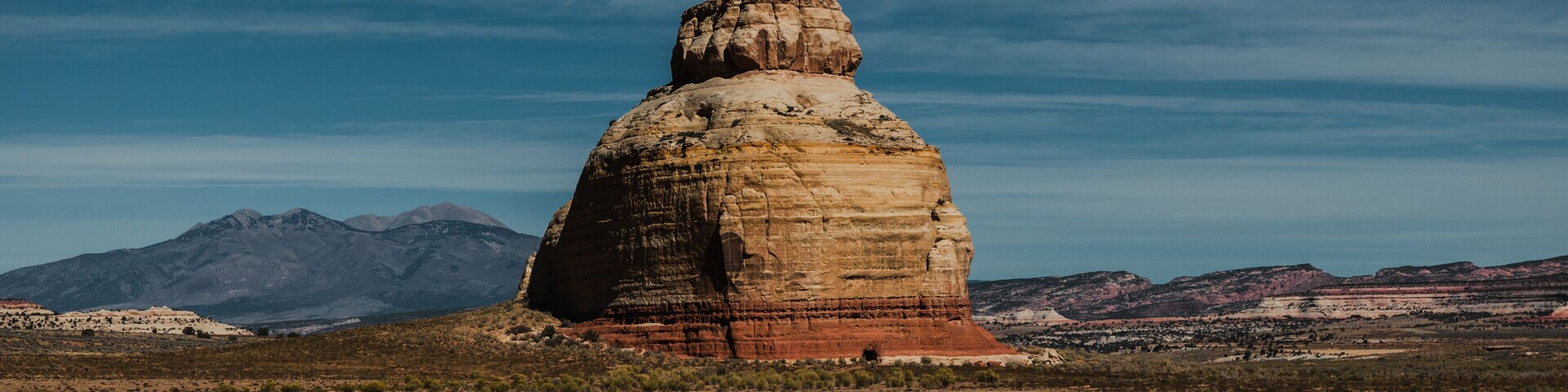 Grande formação rochosa com topo plano e pico menor, rodeada por uma paisagem desértica esparsa e montanhas ao longe.