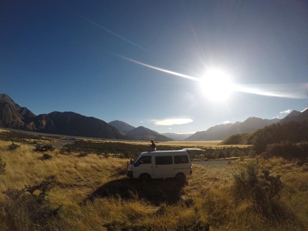 Uma van branca estacionada em um campo gramado, com montanhas ao fundo e o sol brilhando intensamente.