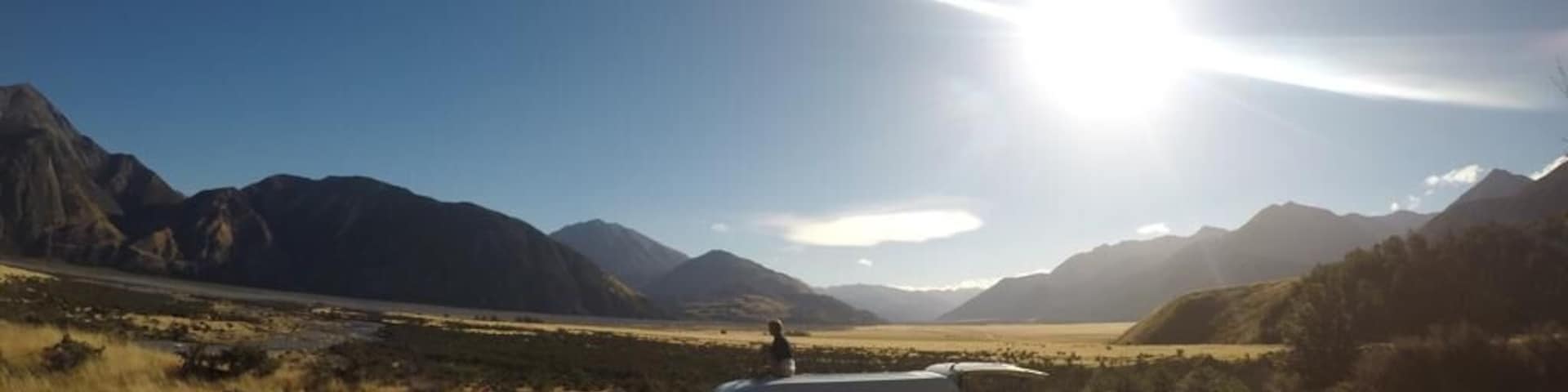 Uma van branca estacionada em um campo gramado, com montanhas ao fundo e o sol brilhando intensamente.