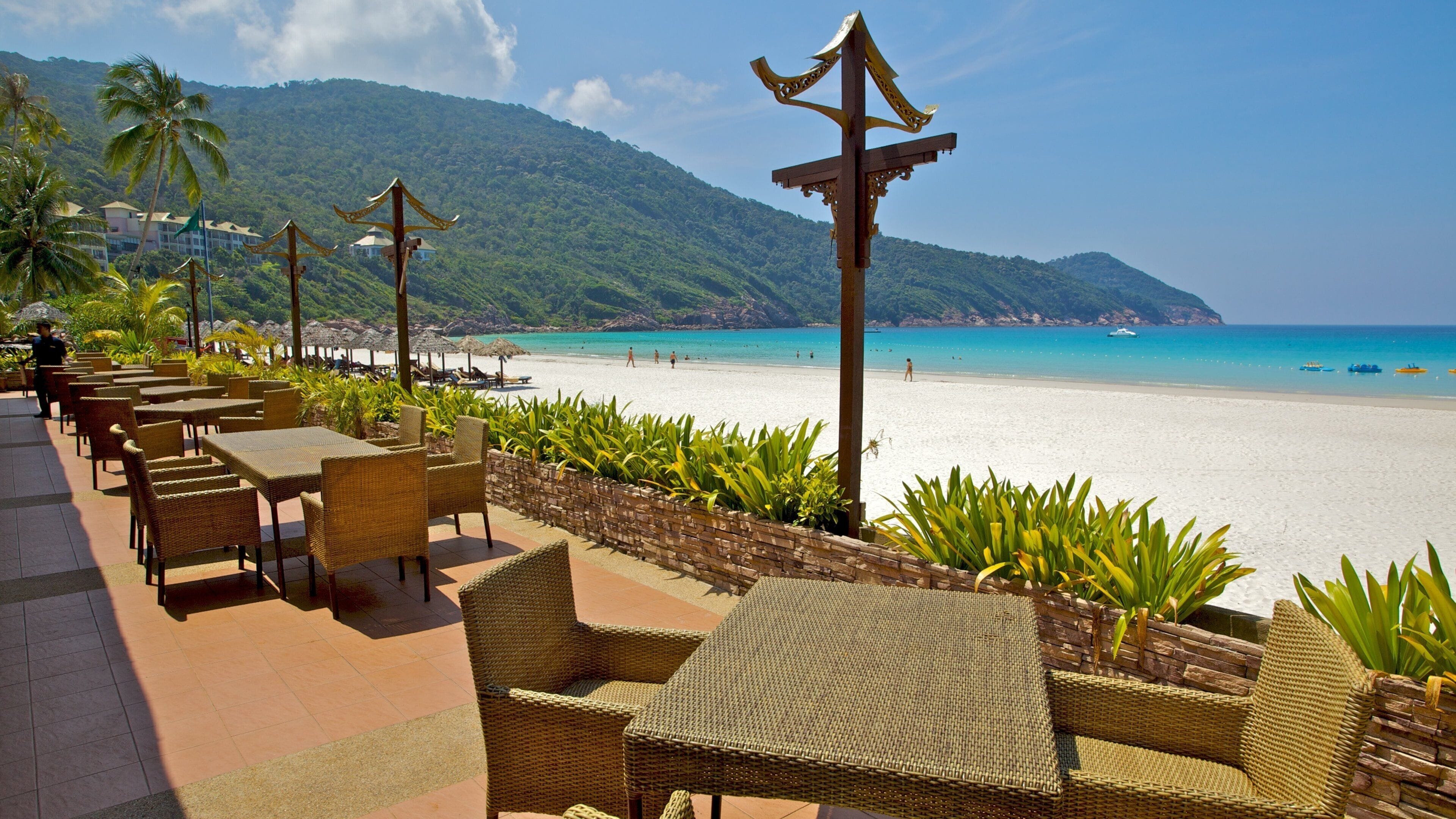 Restaurante à beira-mar com mobiliário de vime, com vista para uma praia arenosa e águas turquesa.