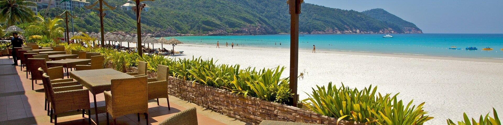 Restaurante à beira-mar com mobiliário de vime, com vista para uma praia arenosa e águas turquesa.