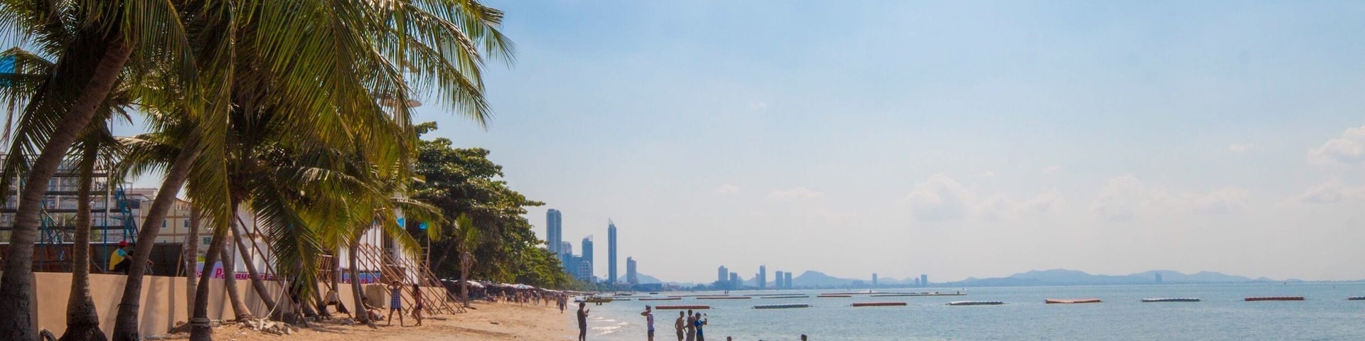 Uma praia arenosa com palmeiras, águas calmas e o skyline da cidade ao longe.