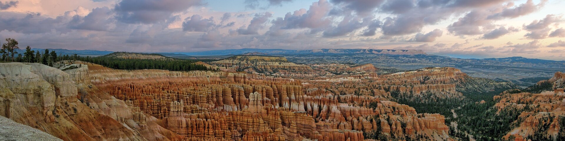 Paisagem acidentada de cânion com formações rochosas em camadas e uma cadeia montanhosa distante sob um céu parcialmente nublado.