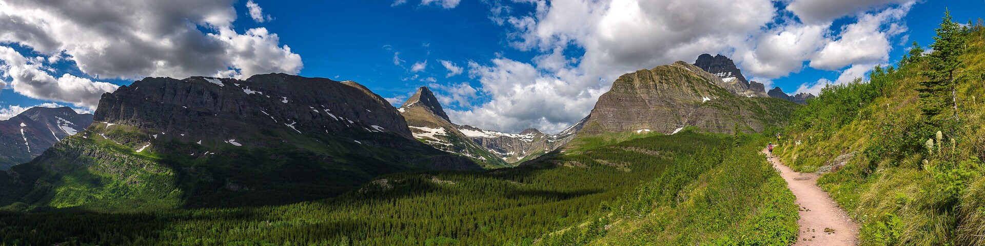 Paisagem montanhosa com trilha de terra, floresta densa e picos nevados sob um céu parcialmente nublado.