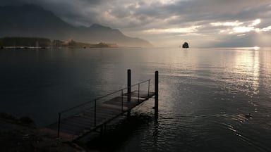 Um lago tranquilo com um píer de madeira, montanhas ao fundo e o sol se pondo no horizonte.