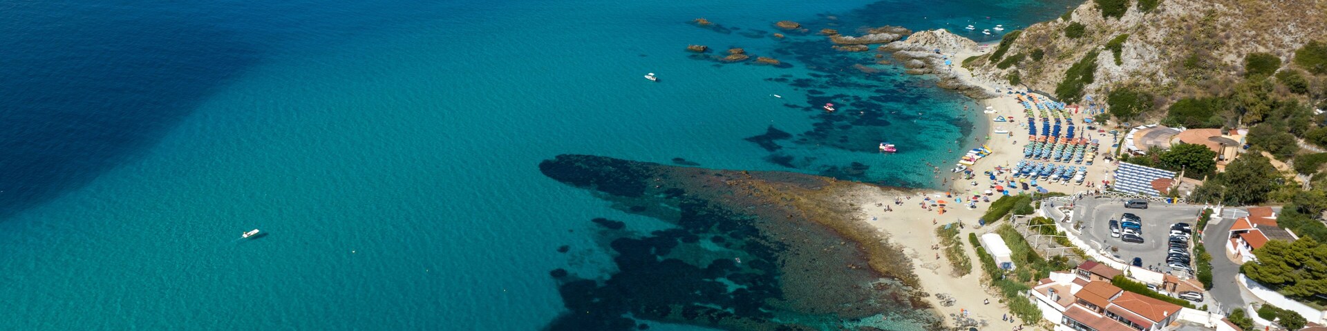 Uma vila costeira com águas turquesa cristalinas, falésias rochosas e uma praia com guarda-sóis e barcos.