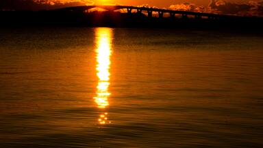 Um pôr do sol sobre águas calmas, com uma ponte em silhueta no céu.