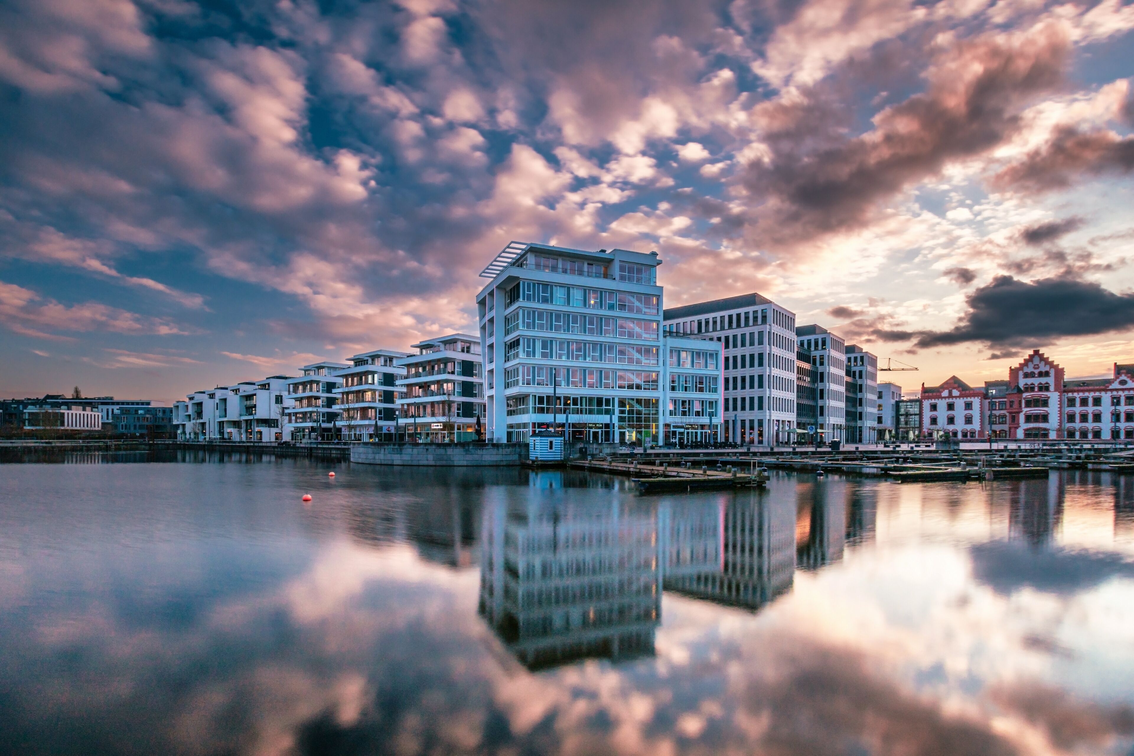 Paisagem urbana à beira-rio com edifícios modernos, um rio calmo e um céu ao pôr do sol.
