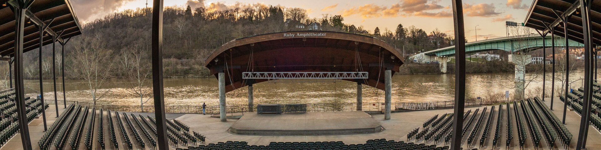 Um anfiteatro com fileiras de assentos verdes, um palco e uma ponte ao fundo.