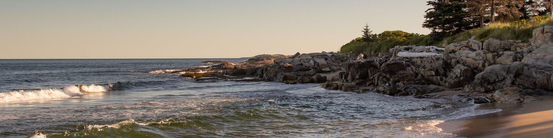 Uma praia com ondas, costa rochosa e árvores.