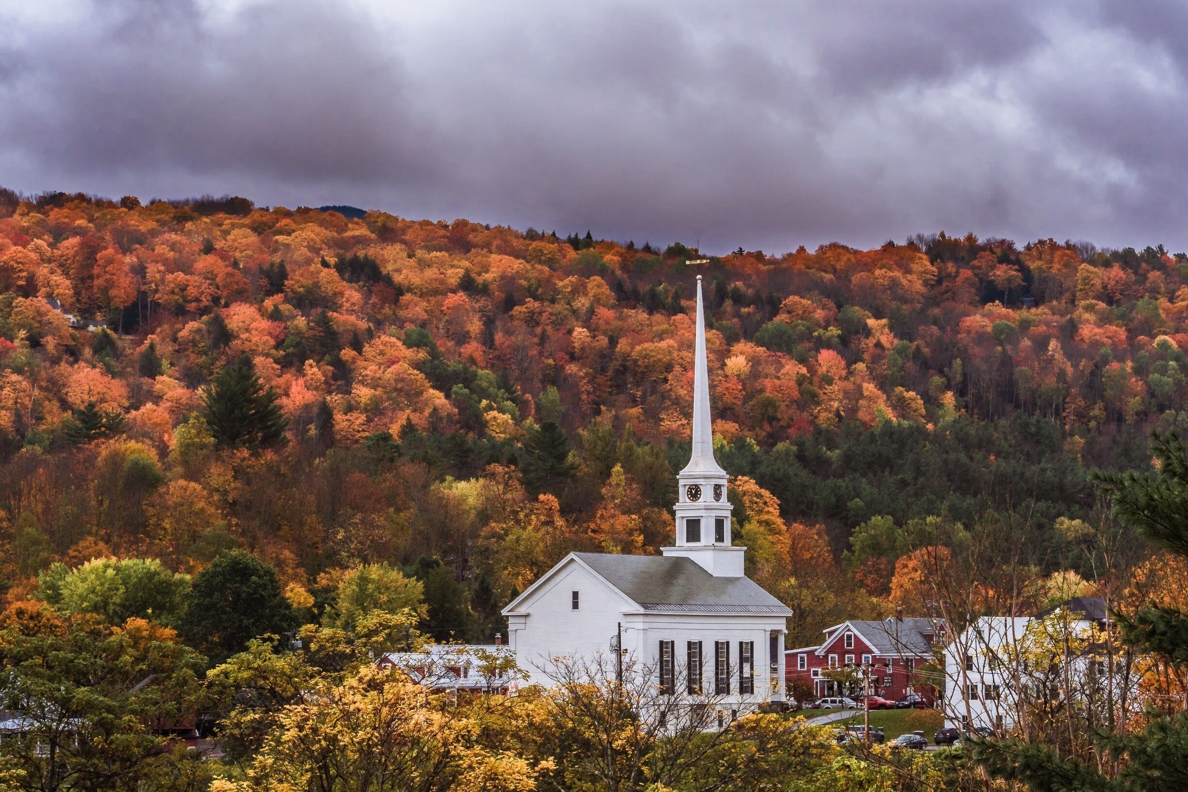 Uma igreja branca com um pináculo e torre do relógio, rodeada por árvores de cores outonais e um céu nublado.