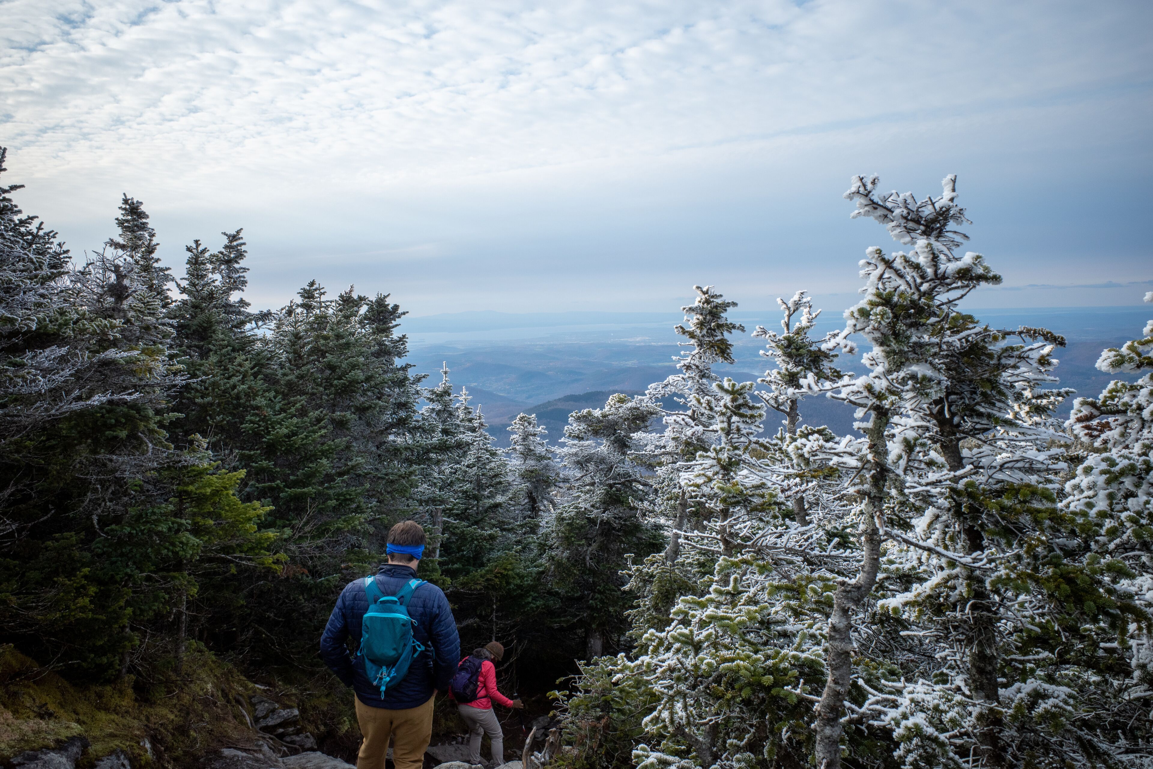 Um caminhante com mochila azul e uma criança com casaco vermelho caminhando por uma floresta nevada, com árvores cobertas de neve e uma cordilheira ao longe.