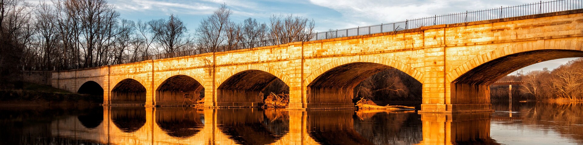 Uma ponte de pedra com várias arcos sobre um rio calmo, rodeada por árvores sem folhas e um céu claro.