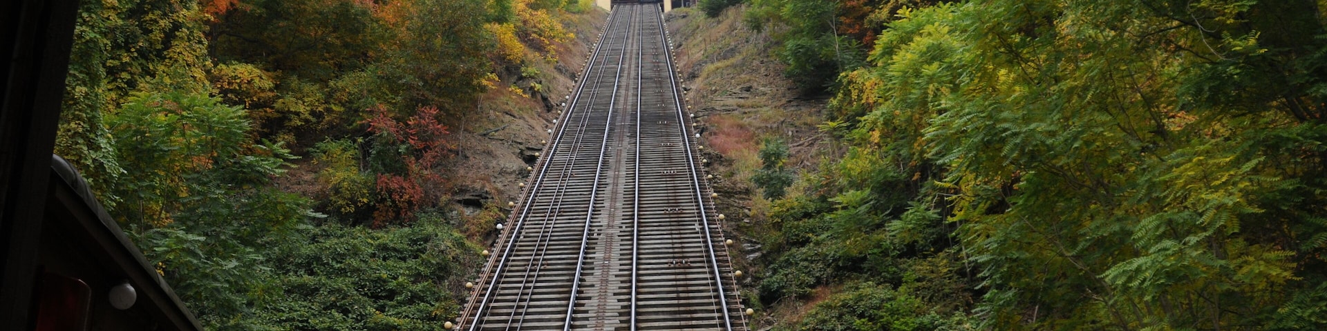 Trilhos de trem subindo por uma área arborizada com árvores de cores outonais.