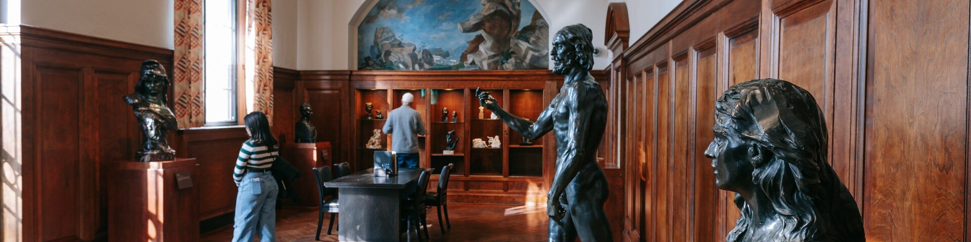 Interior de museu com painéis de madeira, uma pintura grande na parede e esculturas de bronze.