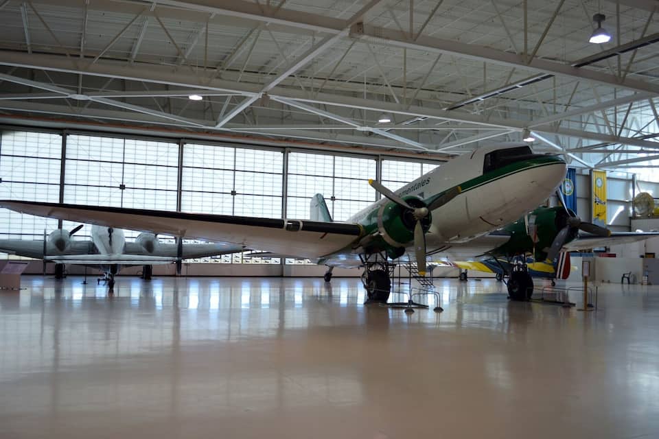 Um hangar com várias aeronaves, incluindo um avião verde e branco e um prateado.
