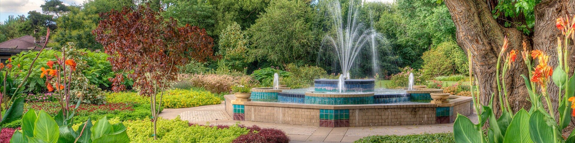 Um jardim com uma fonte, vegetação exuberante e uma variedade de flores coloridas.