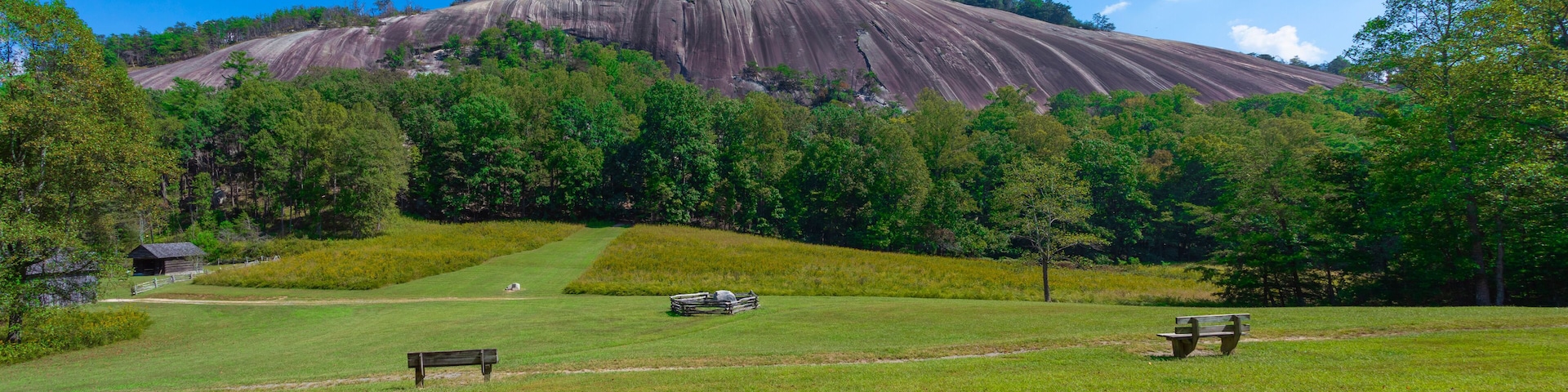 Um campo gramado com uma grande formação rochosa ao fundo, cercado por árvores.