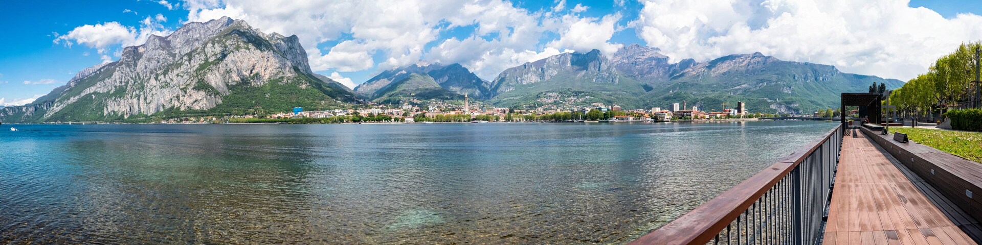 Um lago com uma passarela de madeira, montanhas ao fundo e uma cidade na outra margem.