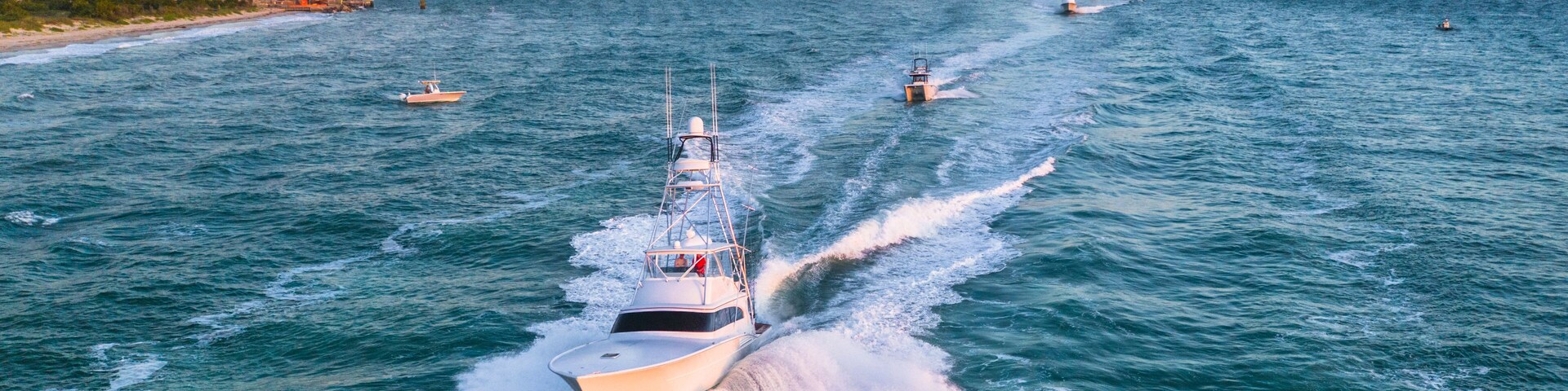 Grande corpo de água com um barco deixando rastro, ladeado por uma costa distante com edifícios e céu claro.