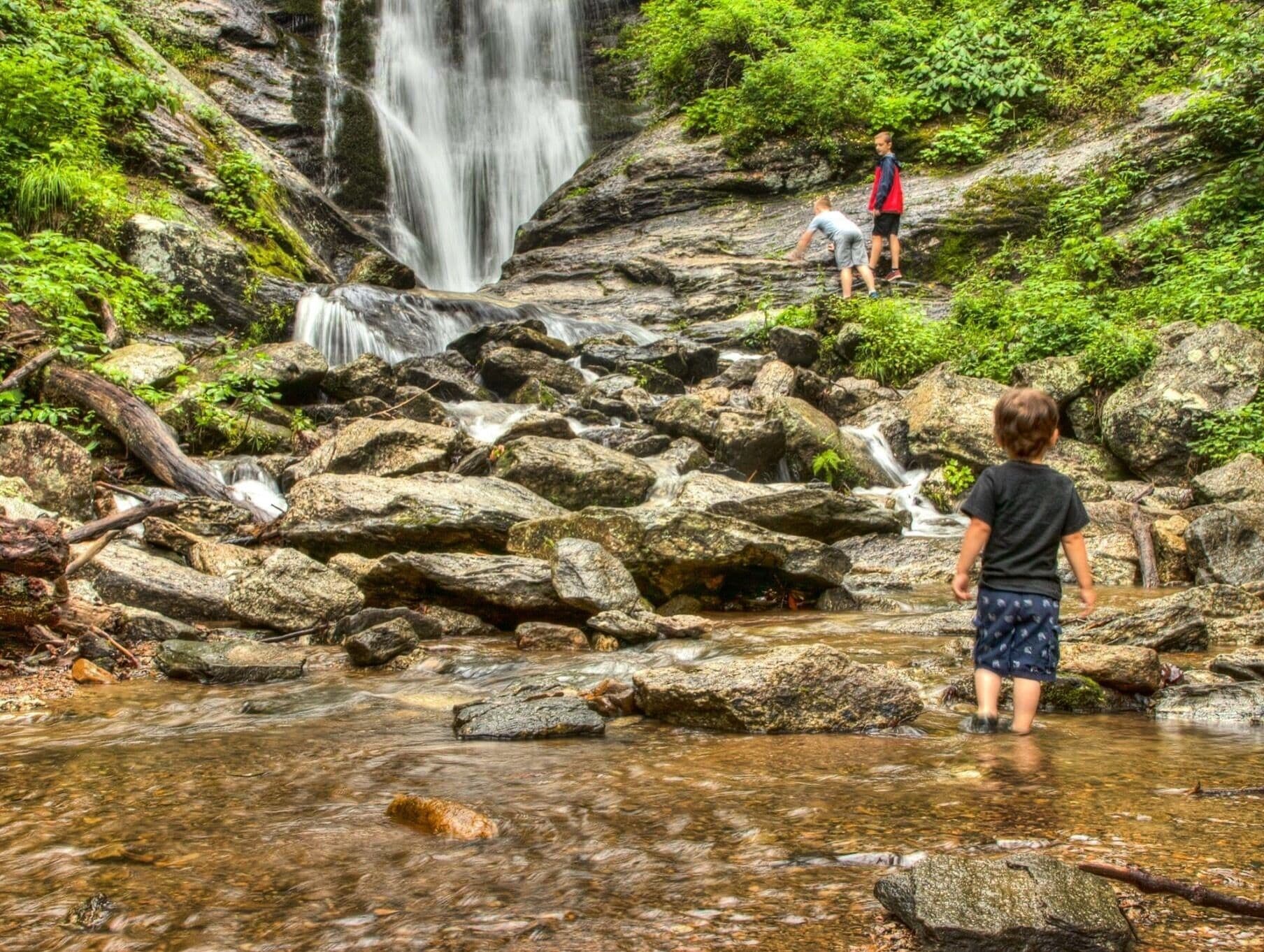 Uma cascata desce sobre rochas em uma área arborizada, com duas pessoas em um afloramento rochoso acima da água.
