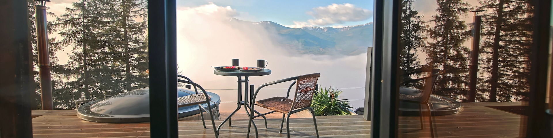 Um deck de madeira com duas cadeiras e uma mesa, com vista para uma paisagem montanhosa com árvores e o céu.