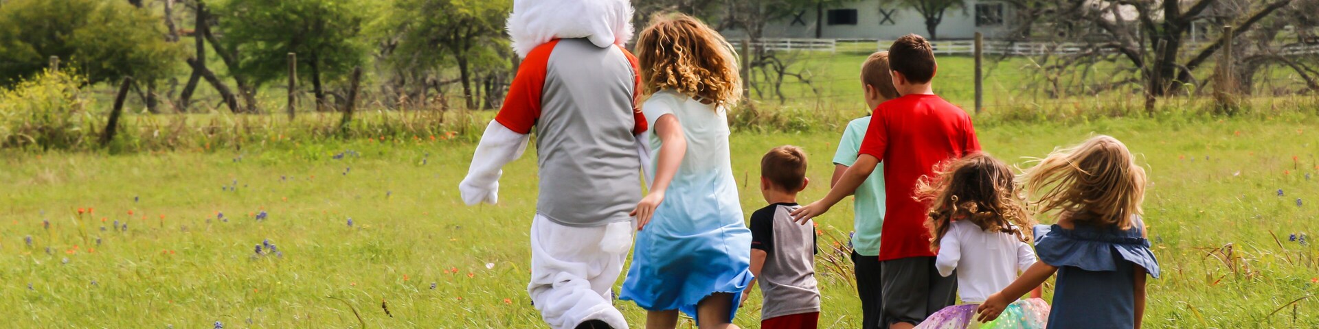 Um grupo de crianças correndo em um campo gramado com uma pessoa vestida de coelho.