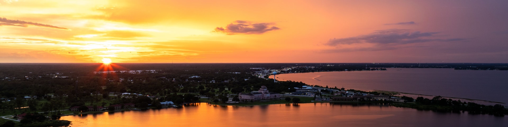 Um pôr do sol sobre um lago calmo, com uma cidade ao primeiro plano e uma ponte que se estende sobre a água.