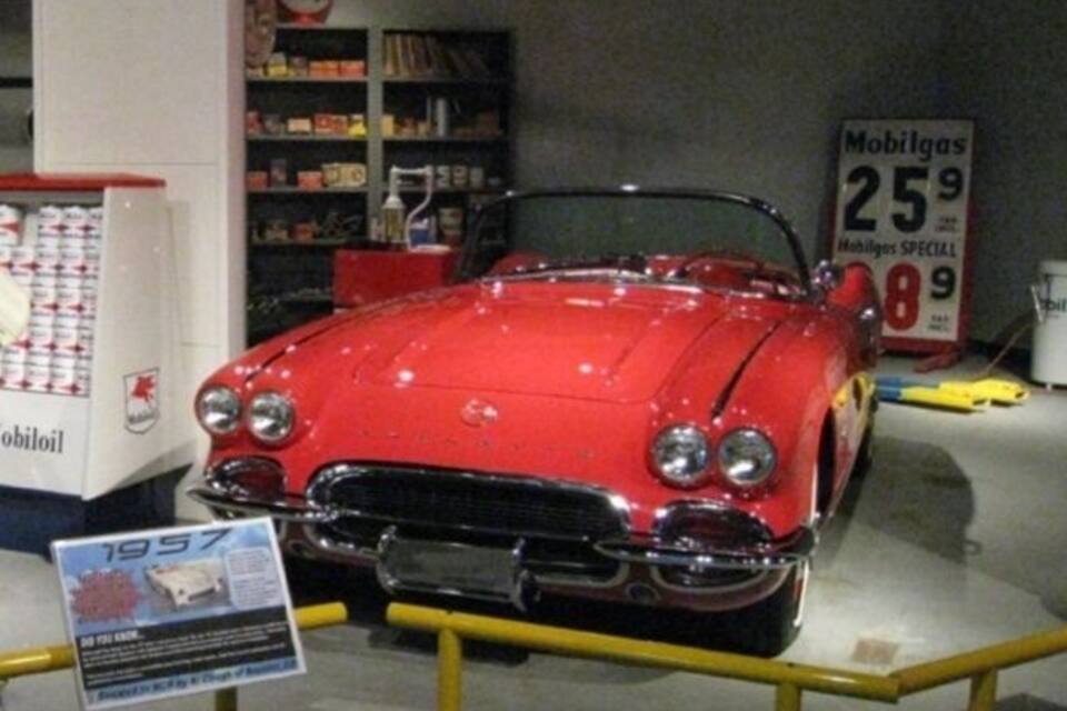 Um carro antigo vermelho em exposição, com um letreiro Mobilgas ao fundo.