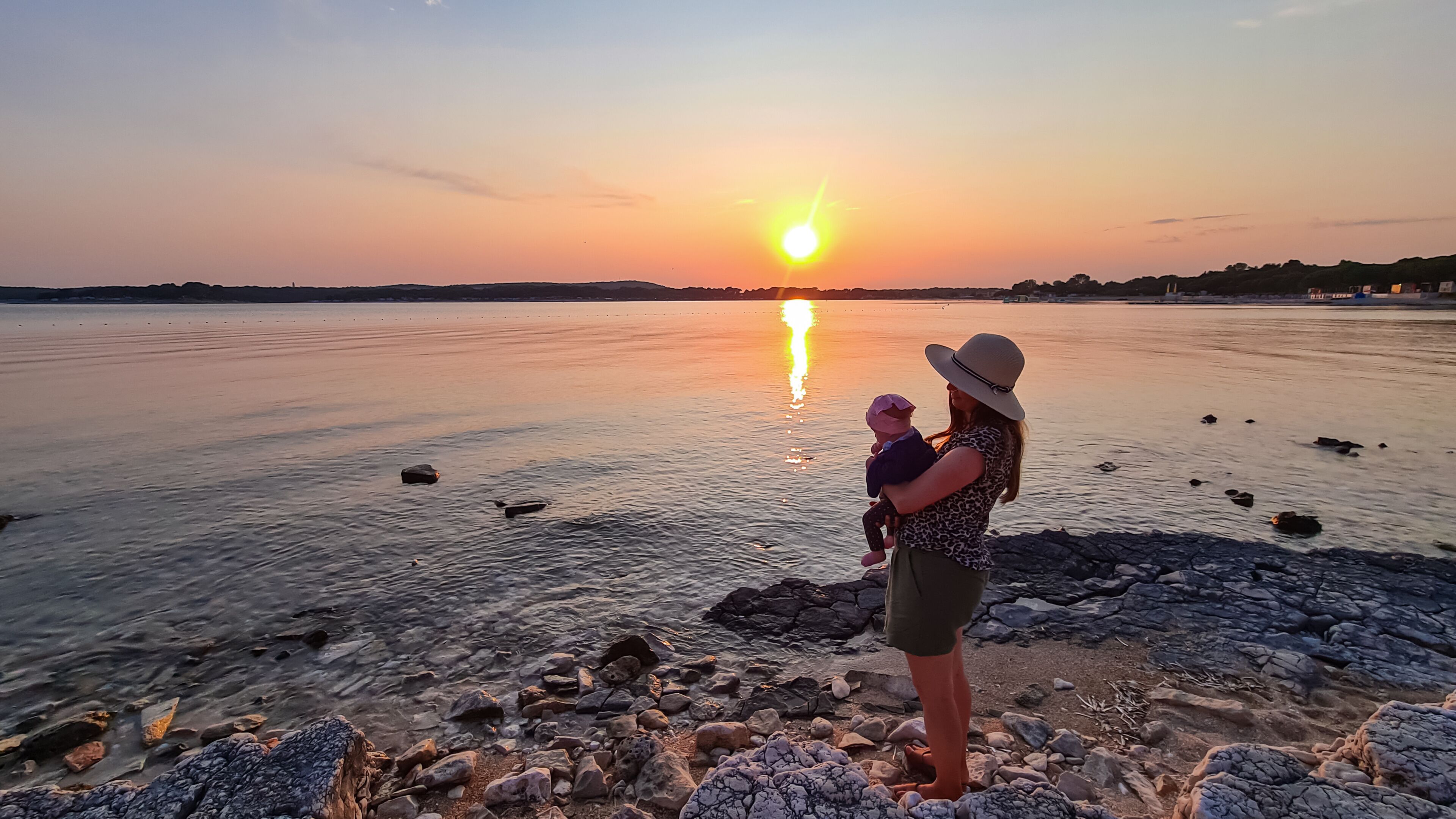Um pôr do sol sobre um lago calmo, com pedras em primeiro plano e uma mulher segurando uma criança.