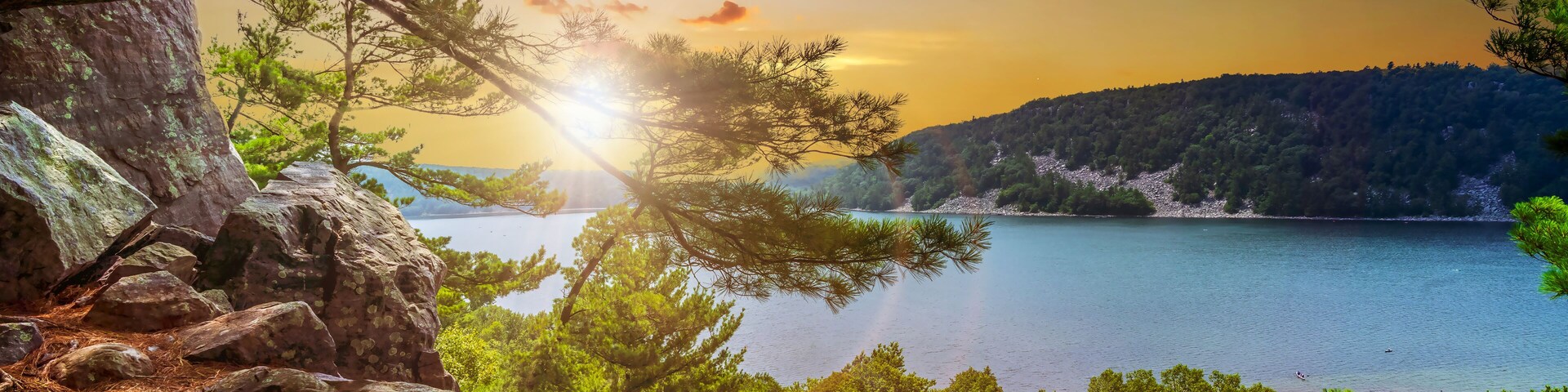 Um lago rodeado por árvores e rochas, com um pôr do sol ao fundo.