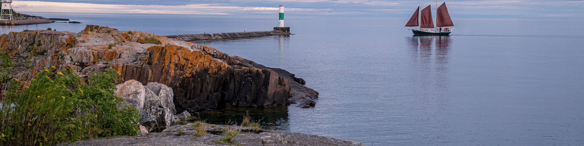 Litoral rochoso com farol e um veleiro na água.