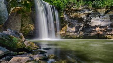Uma cascata deságua sobre rochas em uma piscina, cercada por vegetação.