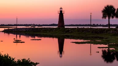 Um farol ao pôr do sol com um lago calmo refletindo a estrutura e palmeiras em primeiro plano.
