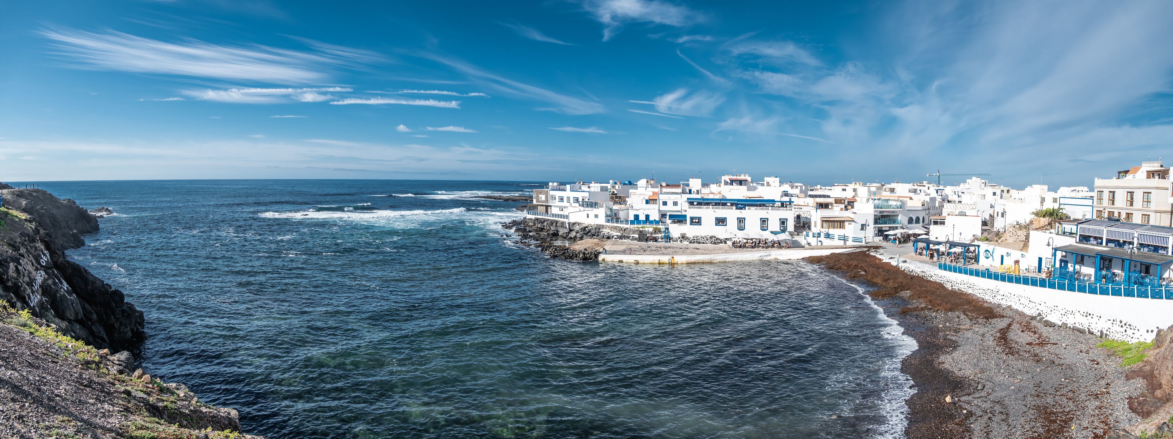 Uma vila costeira com edifícios brancos e detalhes azuis, falésias rochosas e o oceano.