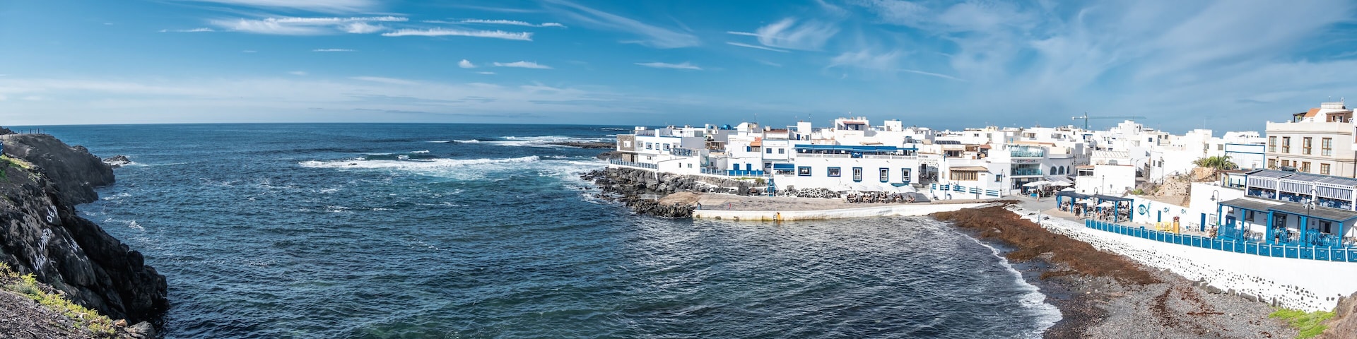 Uma vila costeira com edifícios brancos e detalhes azuis, falésias rochosas e o oceano.