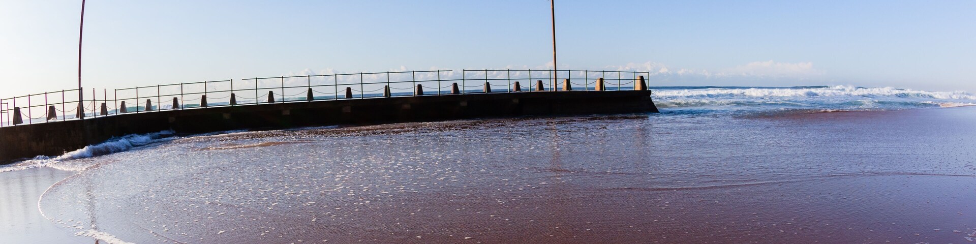 Uma praia arenosa e curvilínea com ondas suaves, um muro de concreto e um corrimão de metal.
