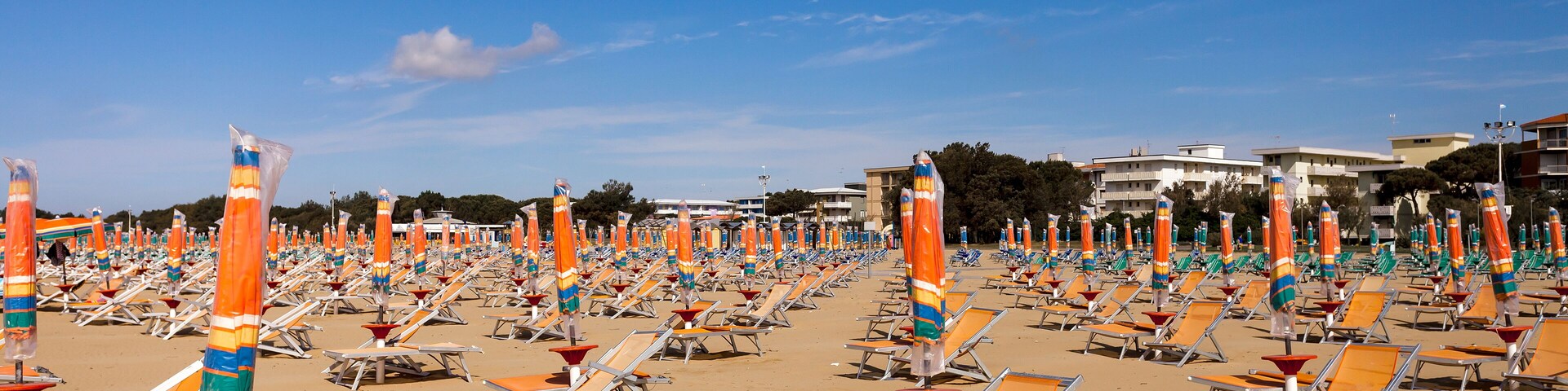 Uma praia com fileiras de espreguiçadeiras e guarda-sóis laranja e branco sob um céu azul claro.