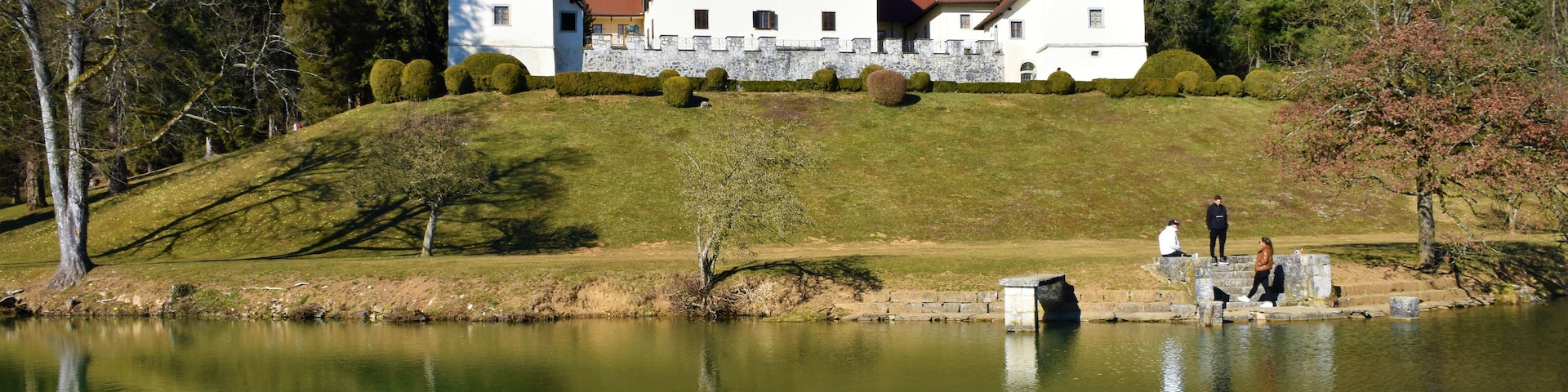 Um edifício grande e branco com telhado vermelho, rodeado por um lago e árvores.