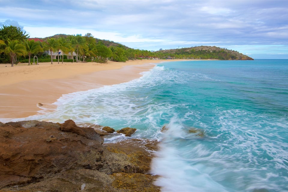 Uma praia com águas turquesa cristalinas, areal arenoso e um promontório rochoso.