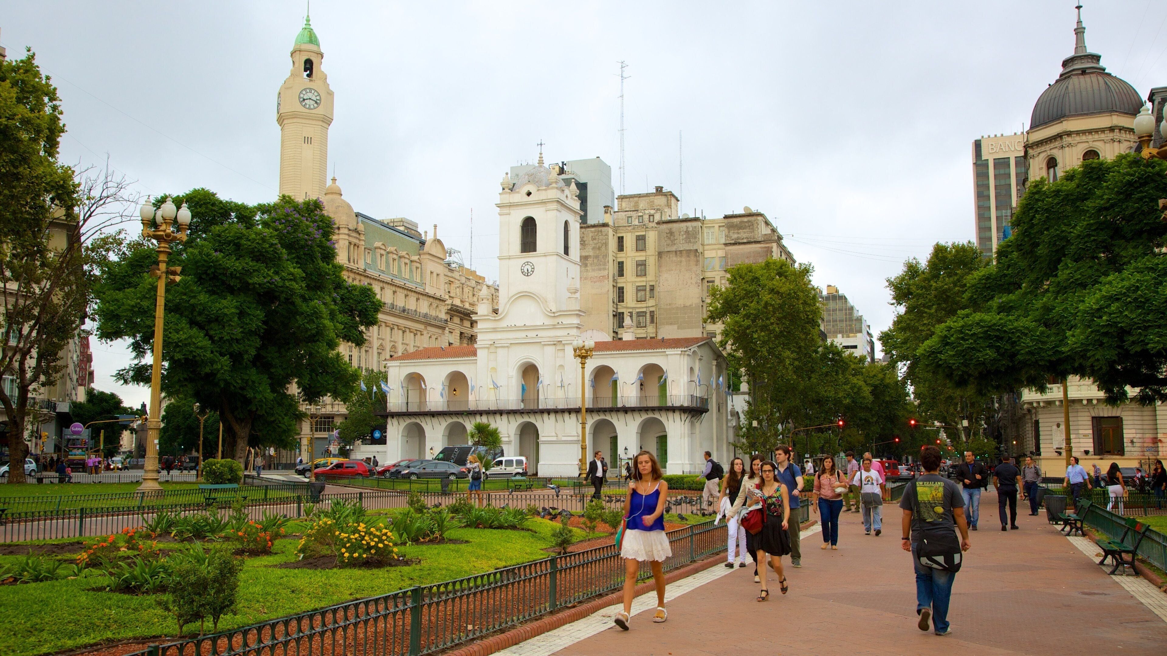 Uma praça histórica com uma imponente torre do relógio, um edifício com cúpula e vegetação exuberante.