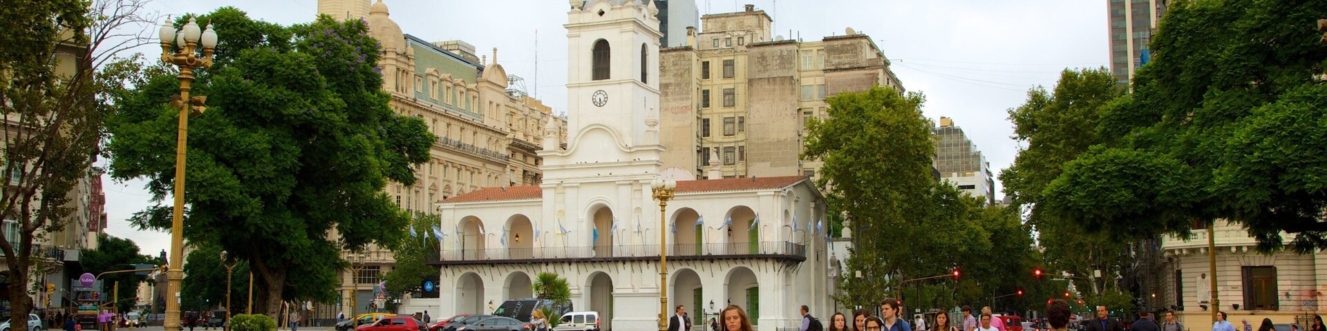 Uma praça histórica com uma imponente torre do relógio, um edifício com cúpula e vegetação exuberante.