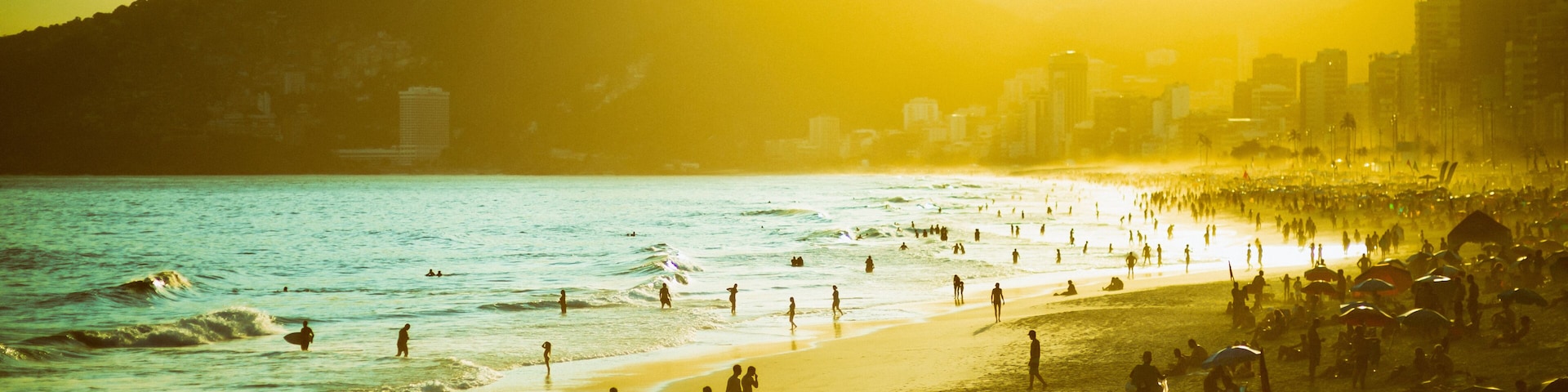 Uma praia cheia de gente, com montanhas e edifícios ao fundo.