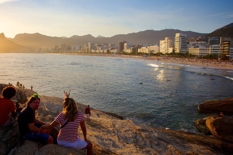 Uma praia com pessoas sentadas em rochas, um horizonte urbano ao fundo e montanhas.