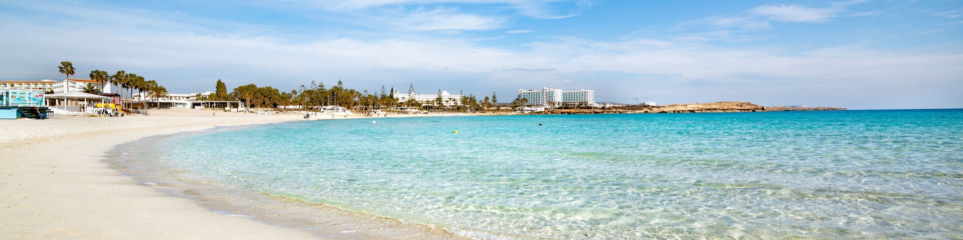 Uma praia arenosa com águas turquesa cristalinas, palmeiras e edifícios ao fundo.