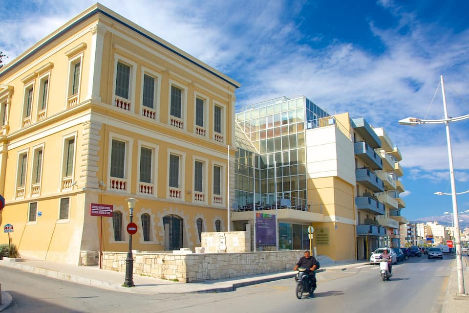 Um edifício histórico amarelo com uma extensão de vidro moderna, uma rua com carros e uma motocicleta, e um céu azul claro.