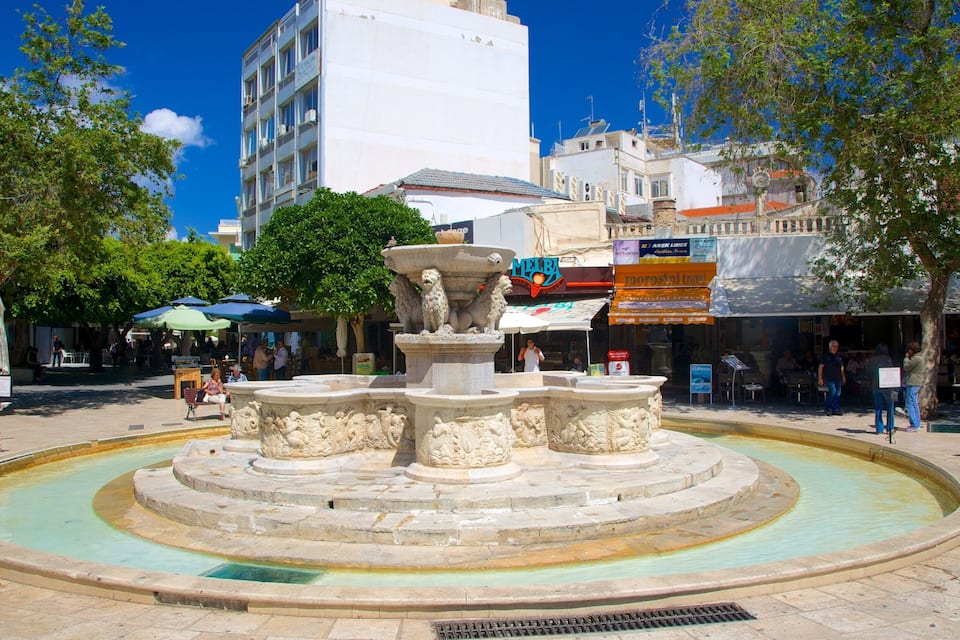 Uma praça pública com uma fonte central, cercada por árvores e edifícios com lojas.